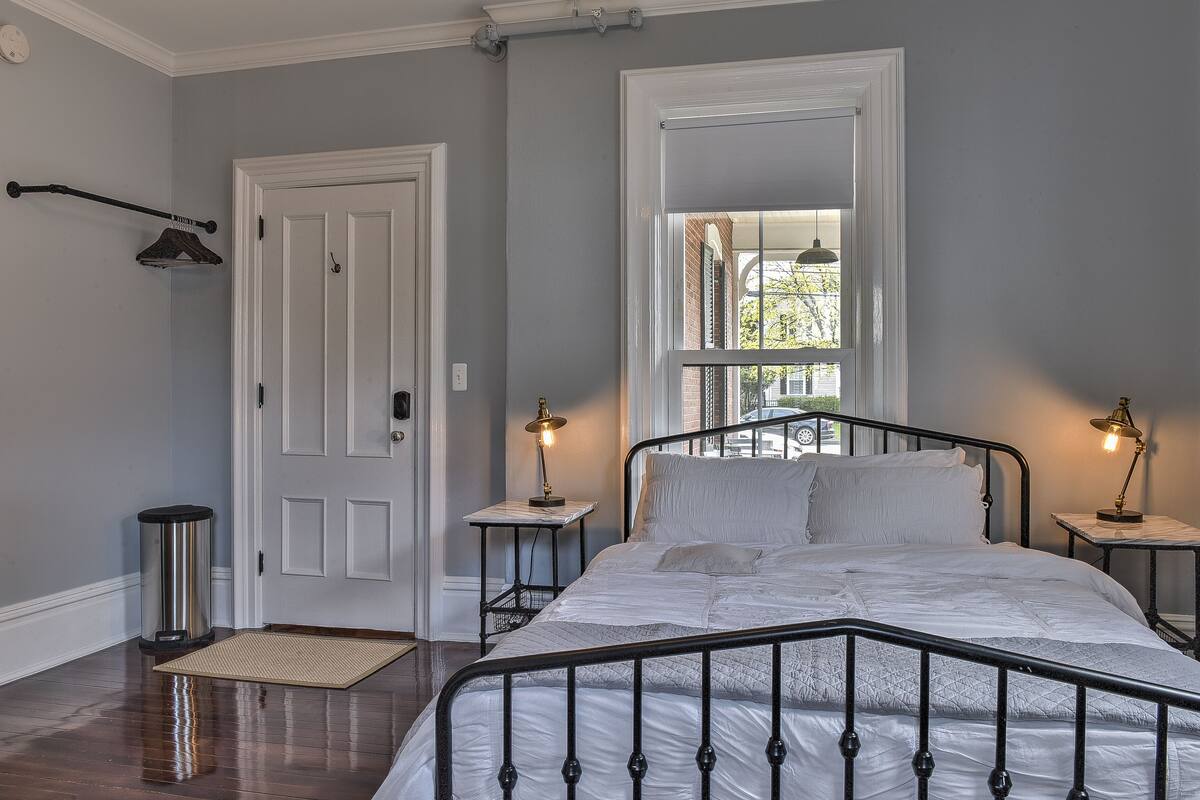 Quarto com cama, mesas de cabeceira, lixeira, porta e janela.