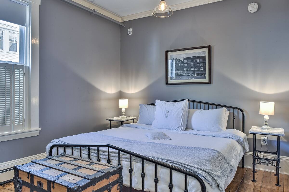 Quarto com estrutura de cama em metal, mesa de cabeceira com luminária, pequena mesa com luminária, quadro na parede e janela com persianas.