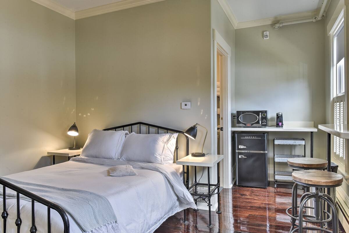Quarto com cama, luminária de cabeceira e pequena cozinha equipada com geladeira, micro-ondas e banquetas.