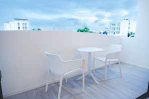 Varanda com duas cadeiras brancas e uma mesa pequena, com vista para uma paisagem urbana com edifícios modernos.