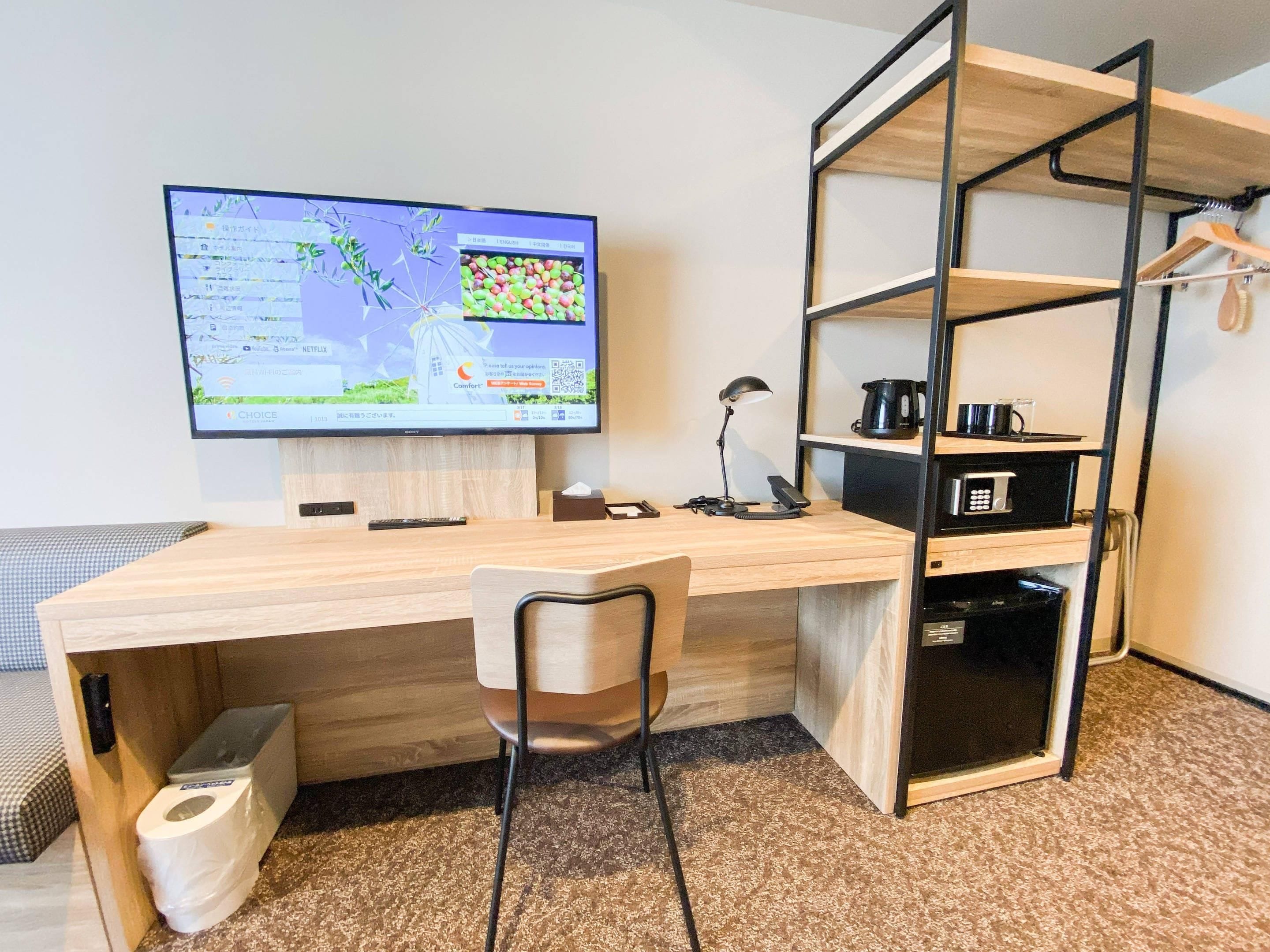 Quarto de hotel compacto com uma secretária de madeira, monitor de computador, cadeira, estante e sanita.