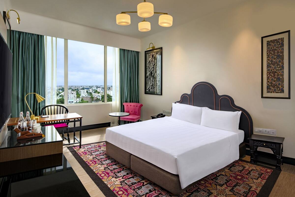 Quarto de hotel com uma cama grande, uma escrivaninha com cadeira, uma mesinha e vista para a cidade por grandes janelas.