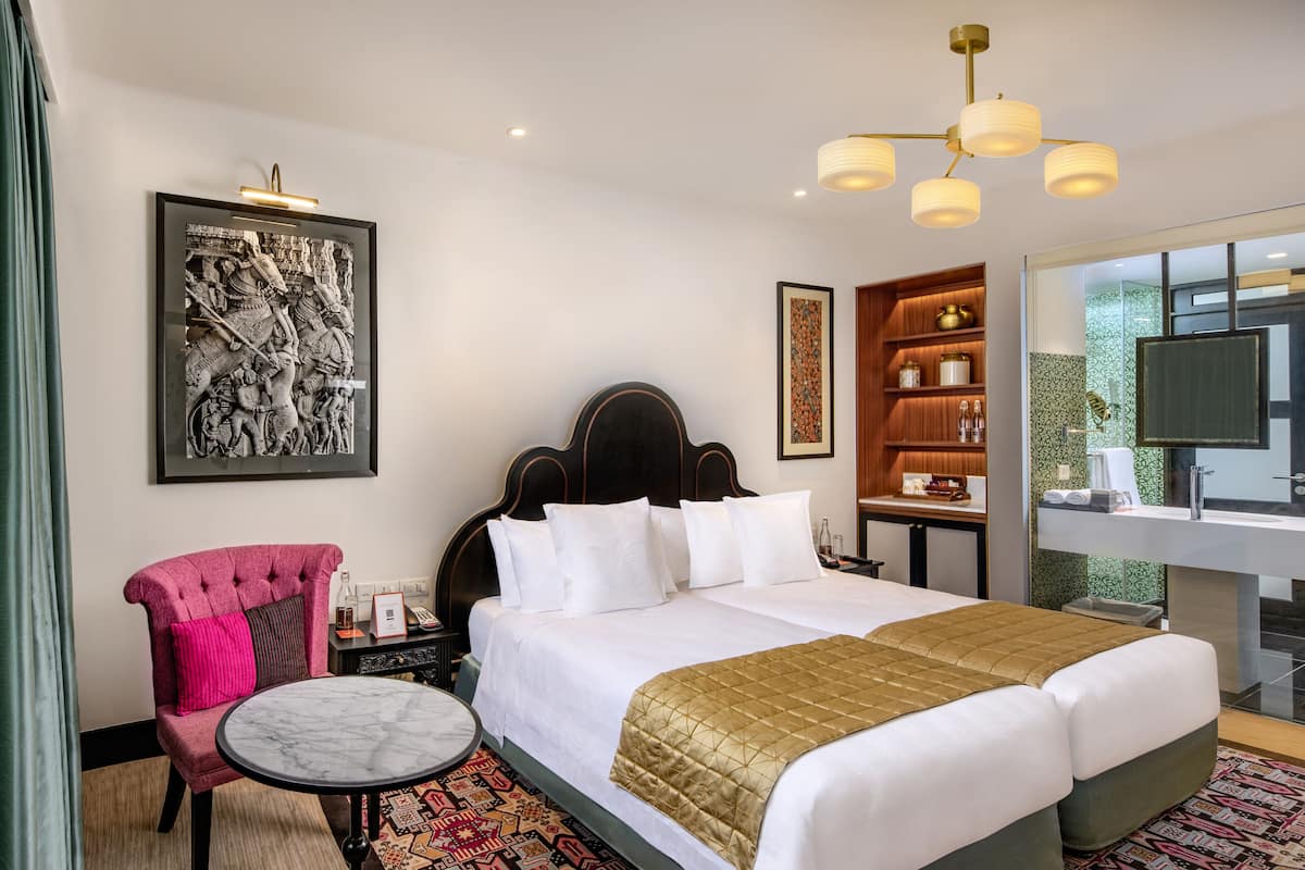 Quarto de hotel com uma cama grande, uma mesa redonda, uma cadeira rosa, um armário de madeira e uma janela ampla.