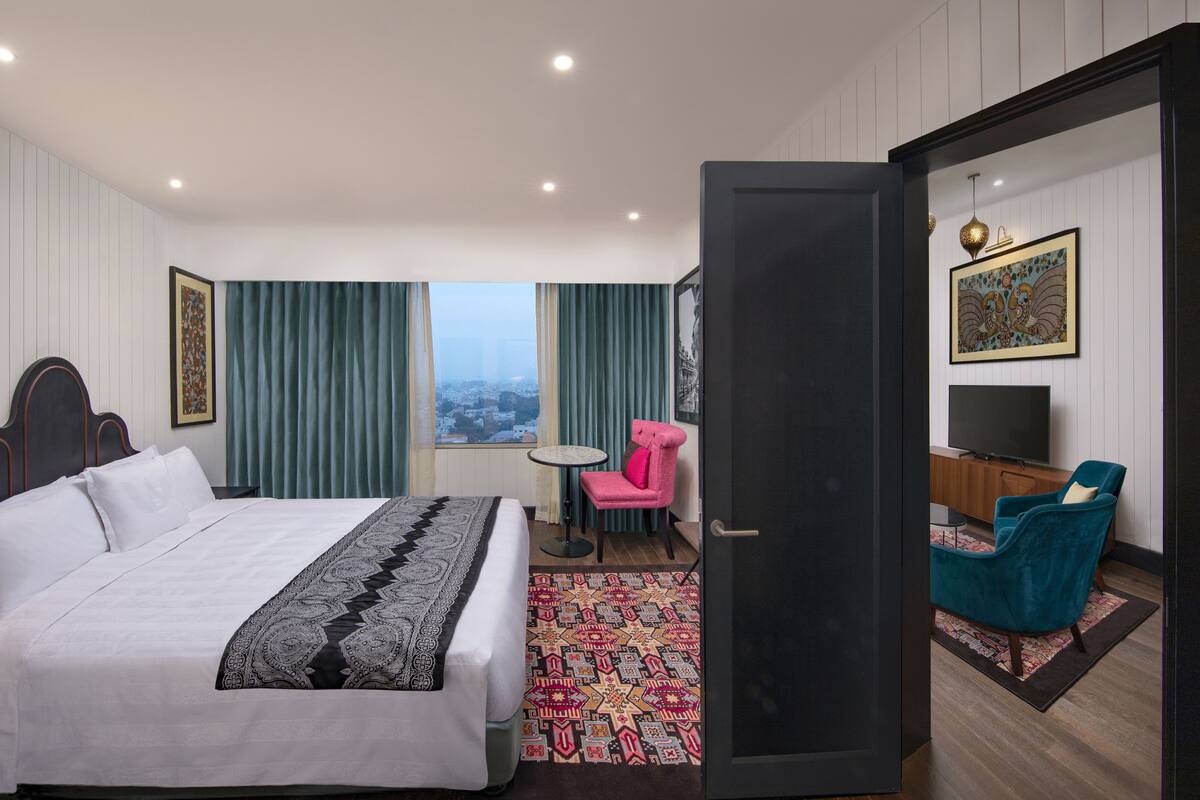 Quarto de hotel com uma cama, uma mesinha, uma cadeira rosa e uma cadeira azul, além de uma televisão.
