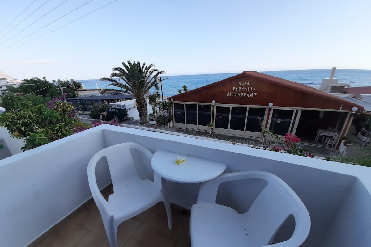 Uma varanda com duas cadeiras de plástico branco e uma mesa pequena, com vista para o restaurante "Agia Roumeli Restaurant" e o mar.