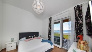 Quarto com cama, mesa de cabeceira, abajur, janela com cortinas e vista para o mar.