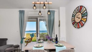 Uma área de jantar com mesa posta para dois, vista para o mar através de grandes janelas e um relógio na parede.
