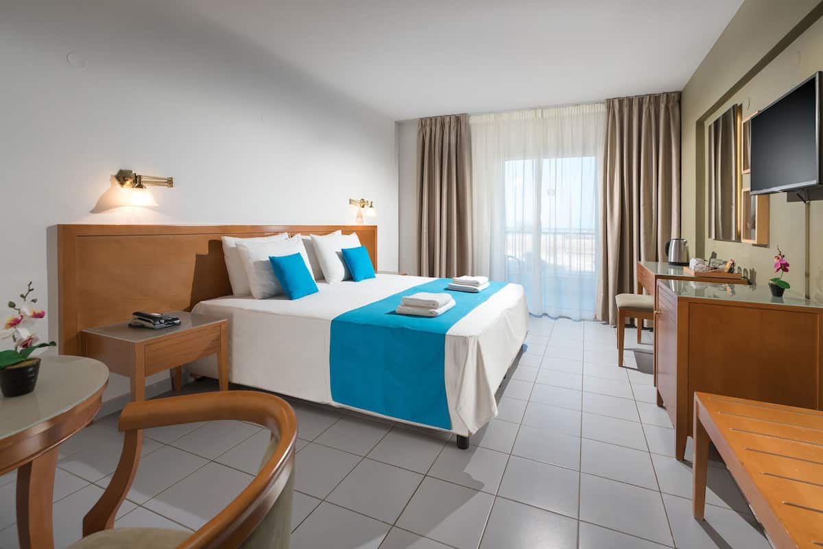 Quarto de hotel com cama, mesas de cabeceira, escrivaninha, cadeira, televisão e janela com cortinas.