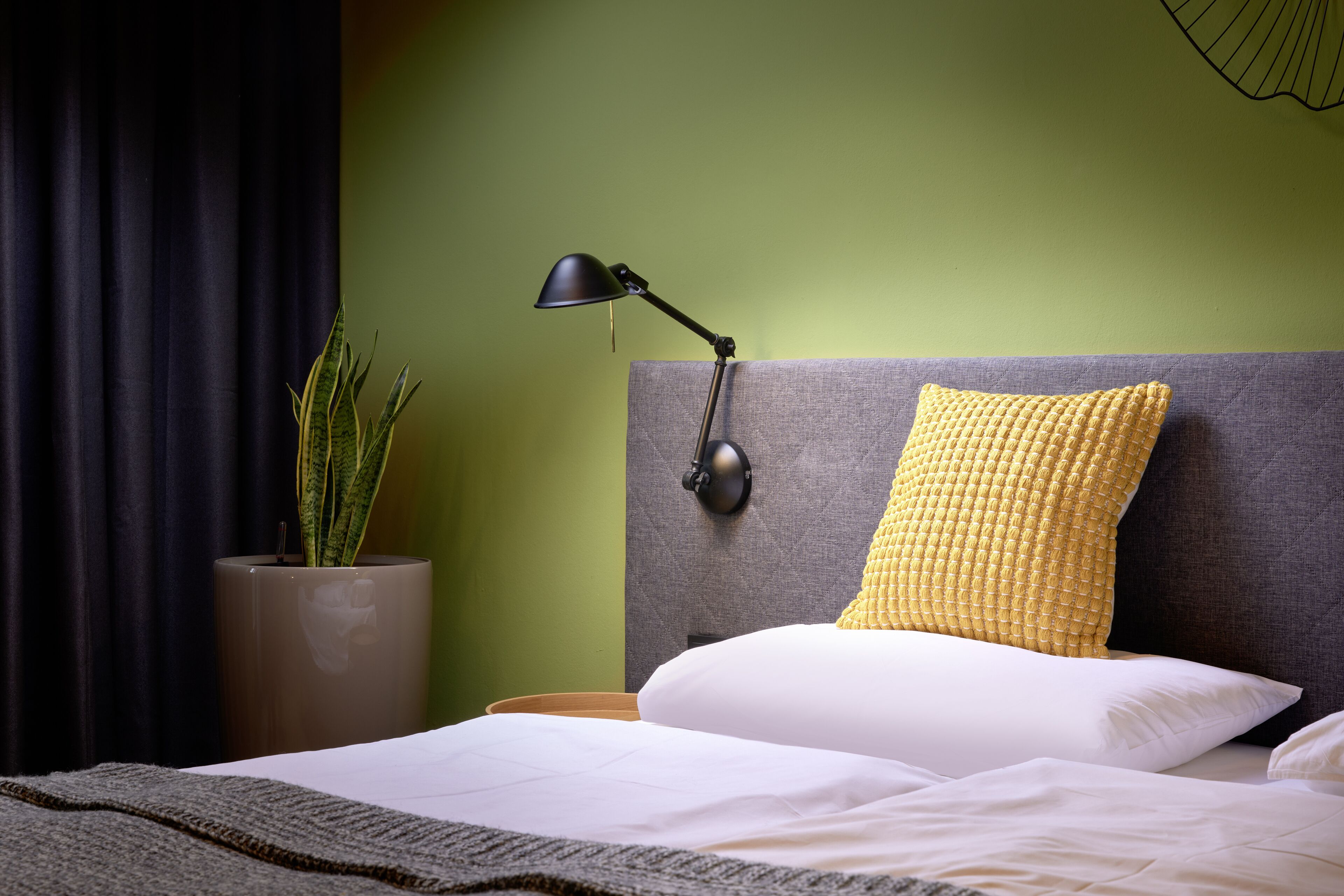 Cama bem arrumada com lençóis brancos, cabeceira cinza e almofada amarela texturizada. Uma luminária de mesa preta está sobre a mesinha de cabeceira, e um vaso com planta é visível no canto.