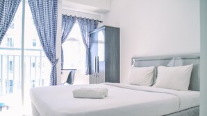 Quarto de hotel moderno com uma cama grande, roupa de cama branca, cabeceira cinza e vista da cidade através de janelas amplas com cortinas estampadas.