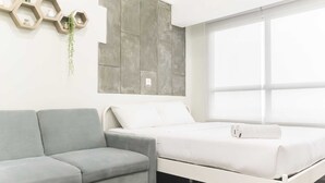 Quarto moderno com um sofá cinzento, uma cama branca e uma janela ampla.