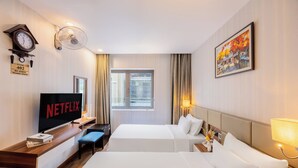 Quarto de hotel com cama, escrivaninha com televisão exibindo 'NETFLIX', relógio, ventilador e um quadro na parede.