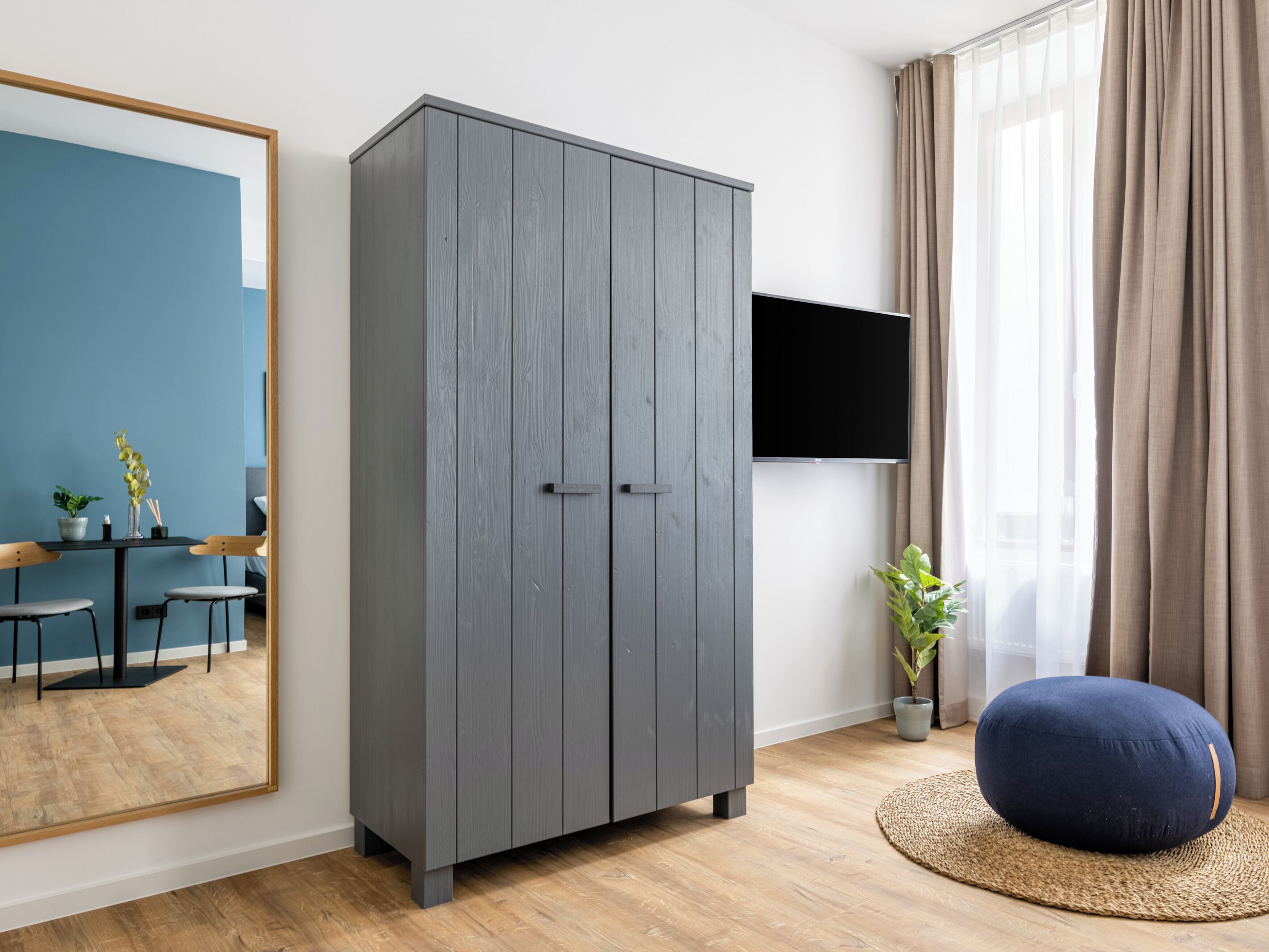 Quarto com guarda-roupa cinzento, televisão na parede, um banquinho redondo e um espelho refletindo uma área de jantar.
