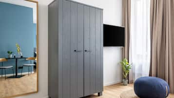 Quarto com guarda-roupa cinzento, televisão na parede, um banquinho redondo e um espelho refletindo uma área de jantar.