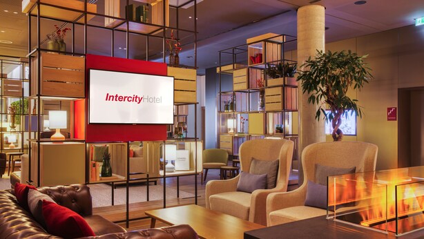 Um lobby de hotel moderno com uma tela grande exibindo o nome do hotel, sofás de couro, poltronas, uma lareira e diversas estantes e plantas decorativas.