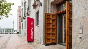Entrada de hotel com placa vermelha, porta de madeira grande e calçada.