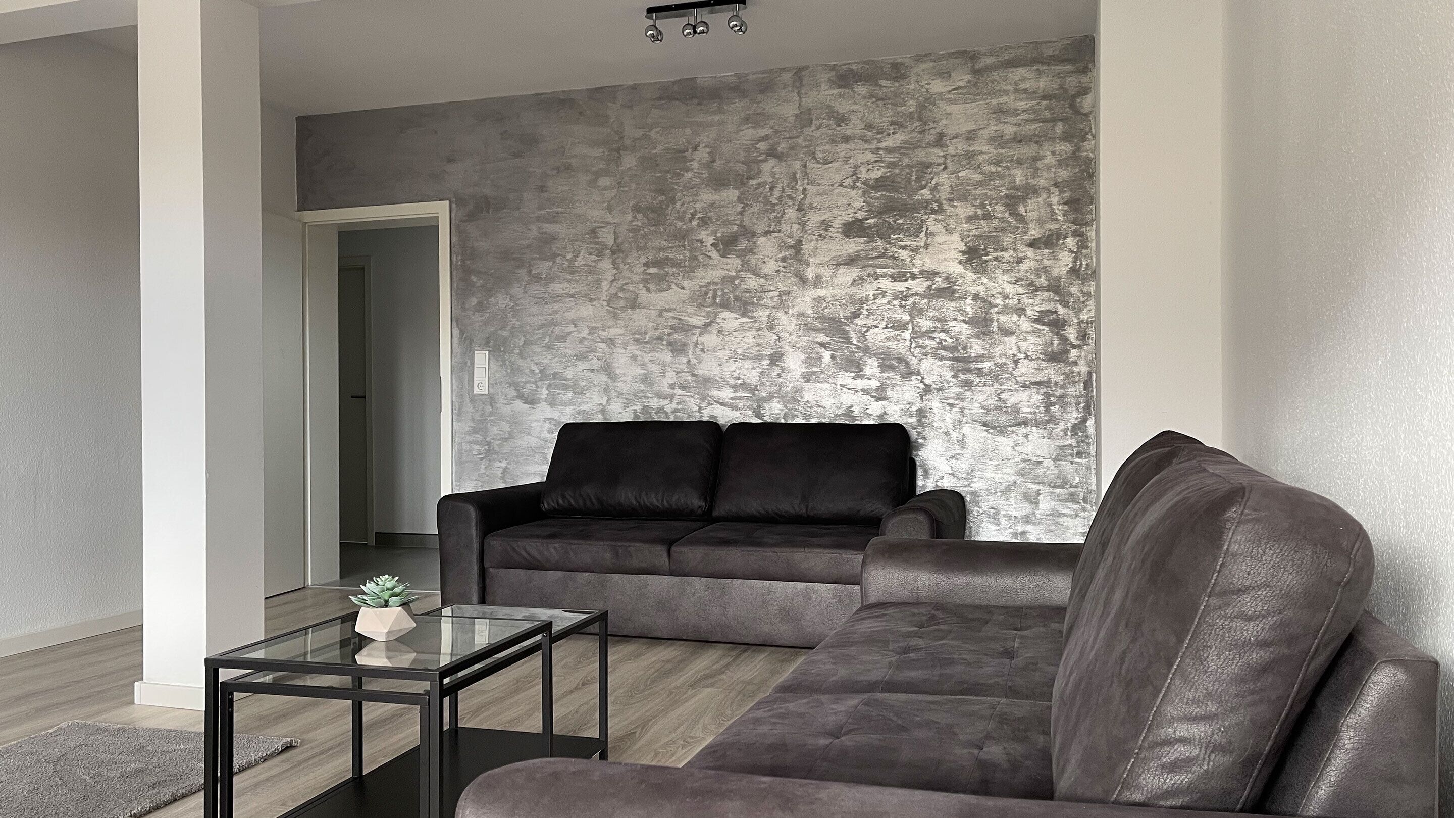 Uma sala de estar moderna com parede texturizada cinza, sofá cinza escuro e mesa de centro de vidro com uma pequena planta.