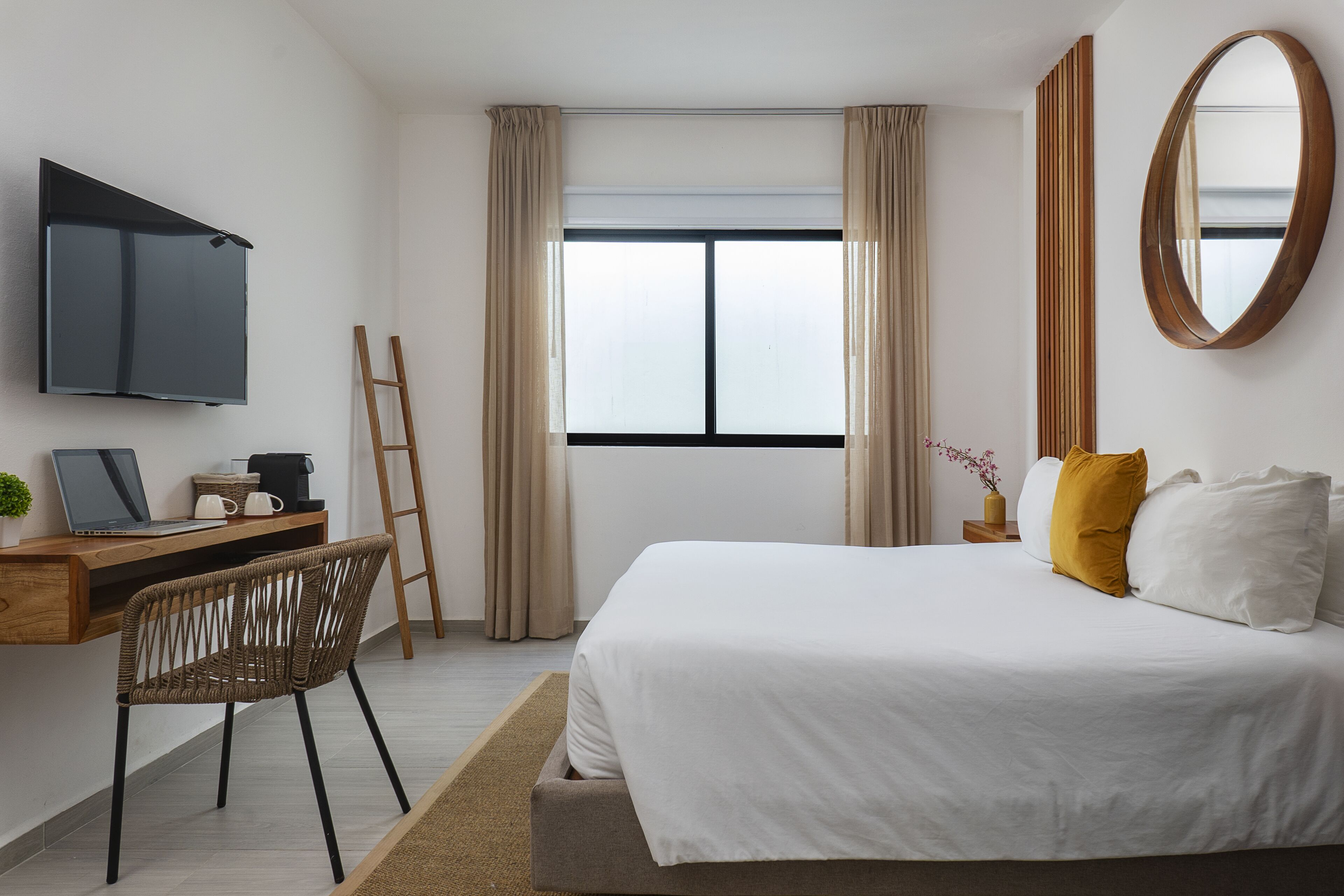 Quarto de hotel moderno com cama, escrivaninha com laptop, cadeira, televisão e janela com cortinas.
