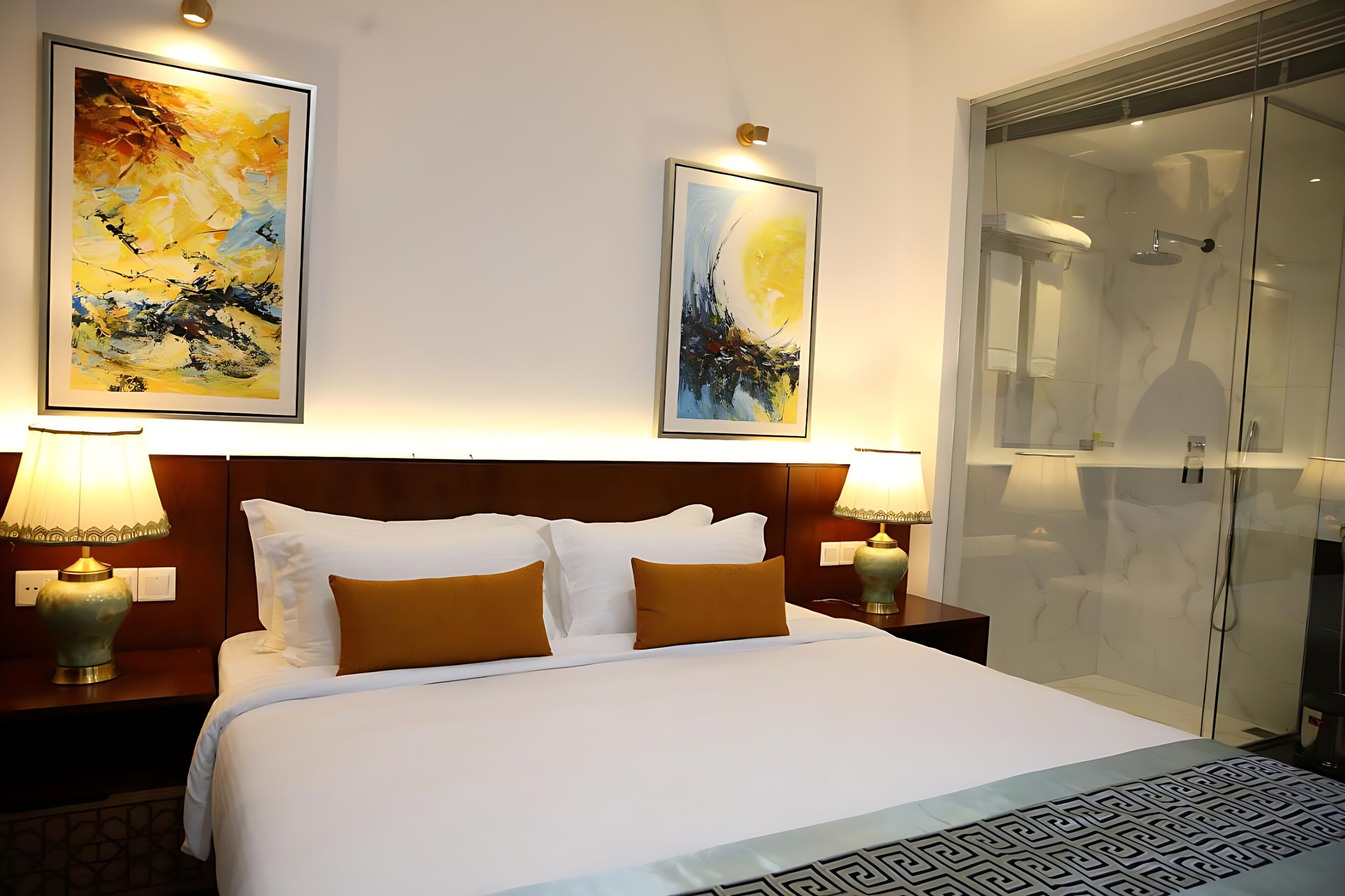 Quarto de hotel com uma cama grande, duas pinturas na parede, um chuveiro com divisória de vidro e duas luminárias de cabeceira.