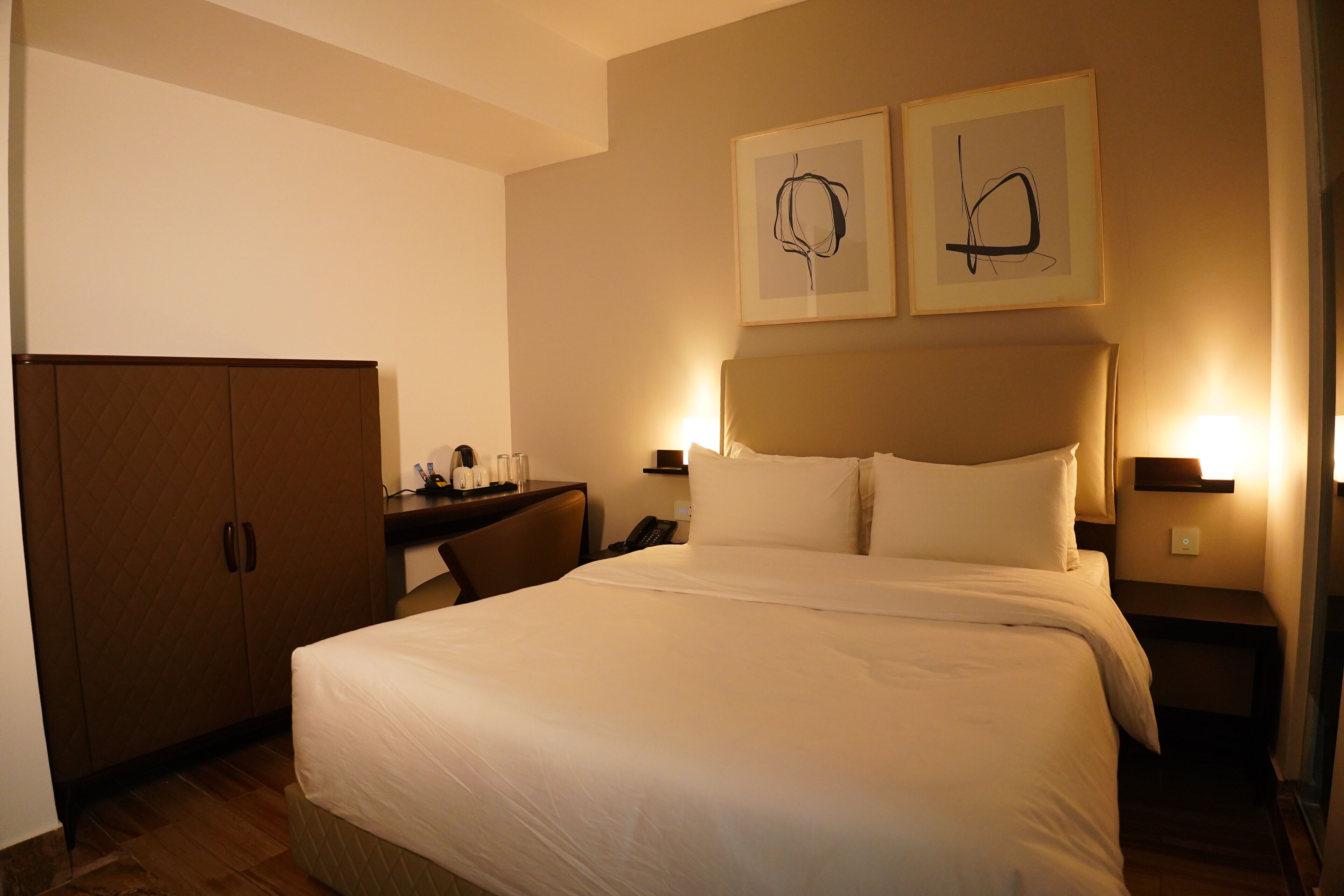 Quarto de hotel com uma cama grande, uma escrivaninha, uma cadeira e duas obras de arte emolduradas na parede.