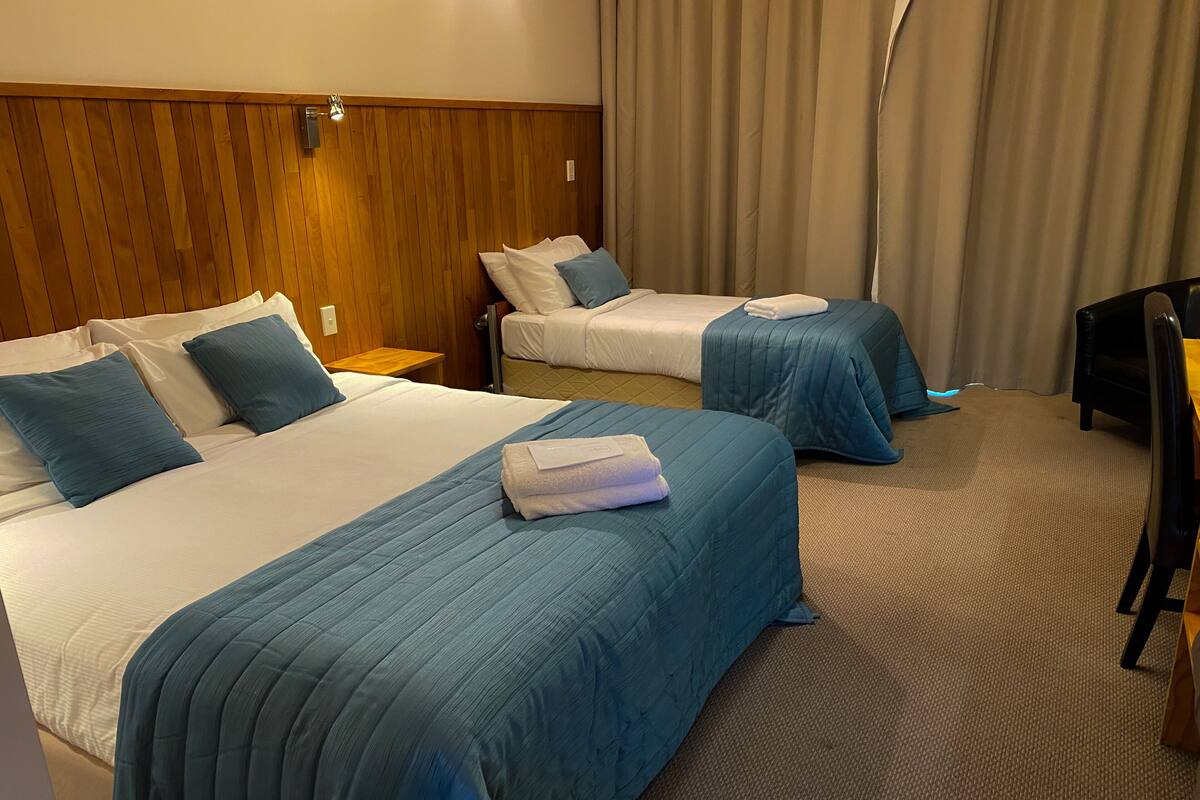 Quarto de hotel com duas camas, cada uma com roupa de cama e almofadas azuis, cabeceira de madeira, mesa de cabeceira pequena de madeira e uma cadeira.