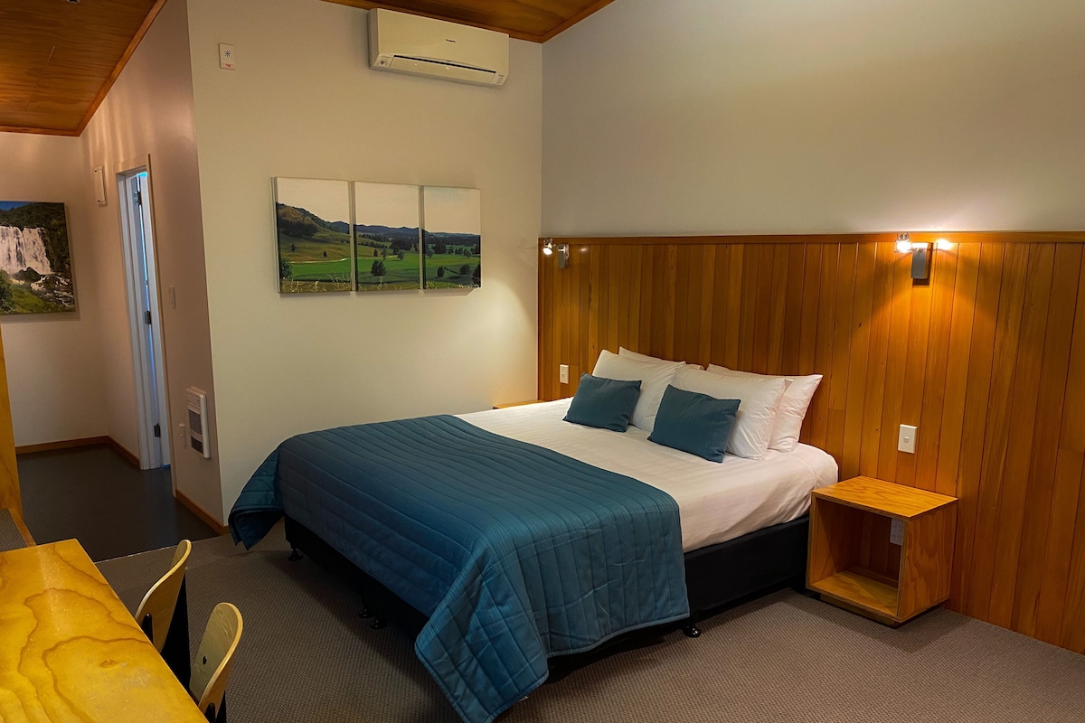 Quarto com cama, mesa de cabeceira, cabeceira de madeira, ar-condicionado fixado na parede e um quadro de uma paisagem.