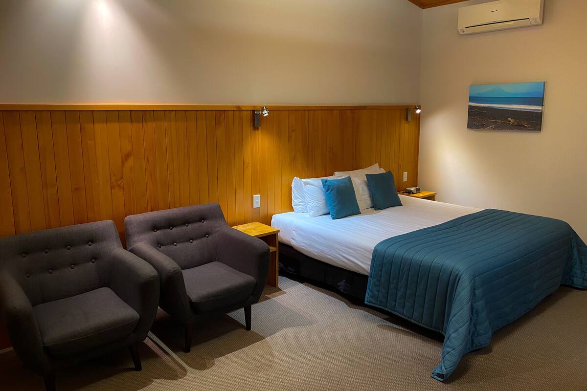 Quarto de hotel com cabeceira de madeira, cama com manta azul, duas poltronas, mesa de cabeceira e um quadro com uma cena de praia.