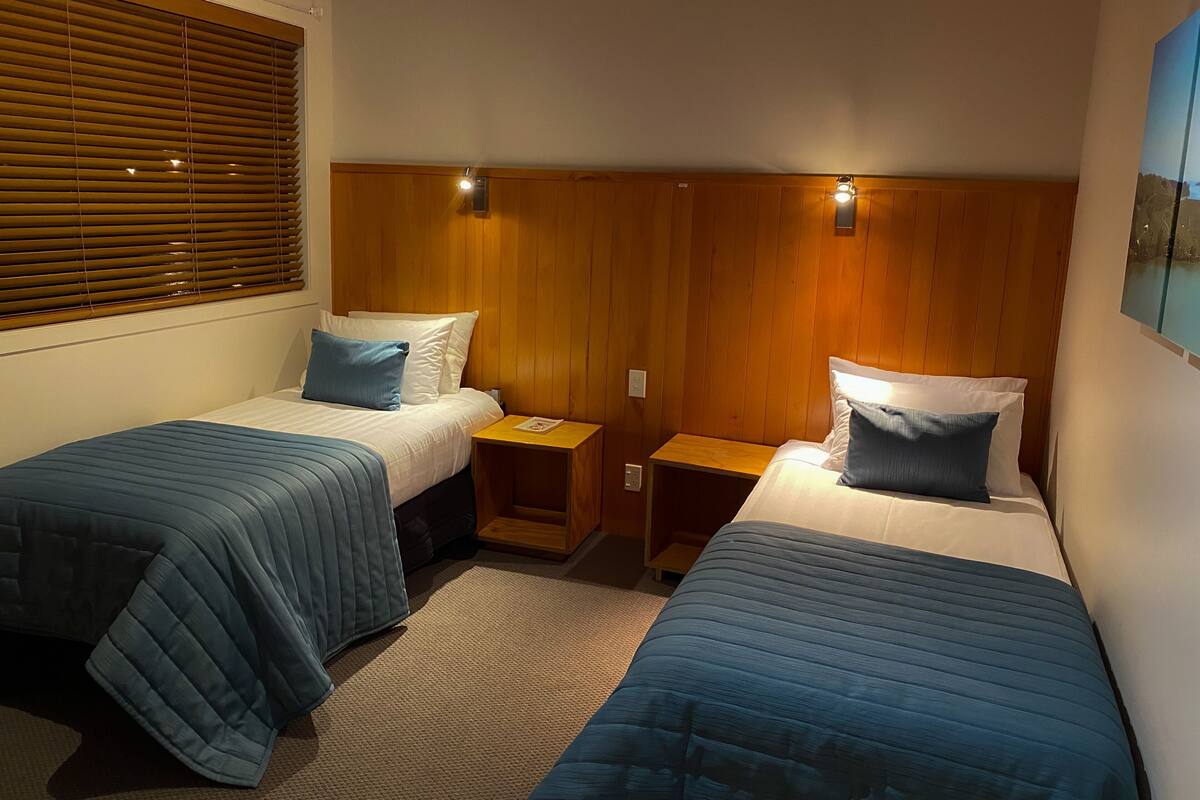 Quarto de hotel com duas camas de solteiro, cabeceiras de madeira, um criado-mudo de madeira pequeno e uma janela com persianas.