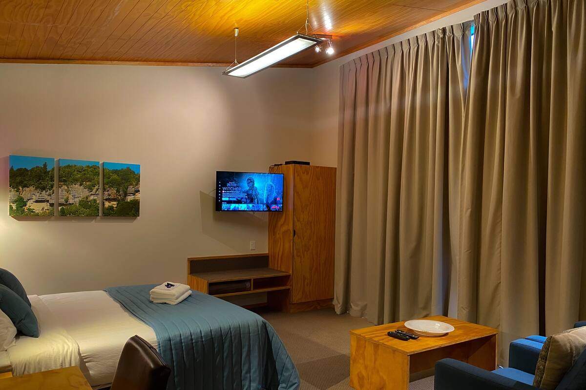 Quarto de hotel com uma cama, televisão presa na parede, armário de madeira, mesinha e sofá.