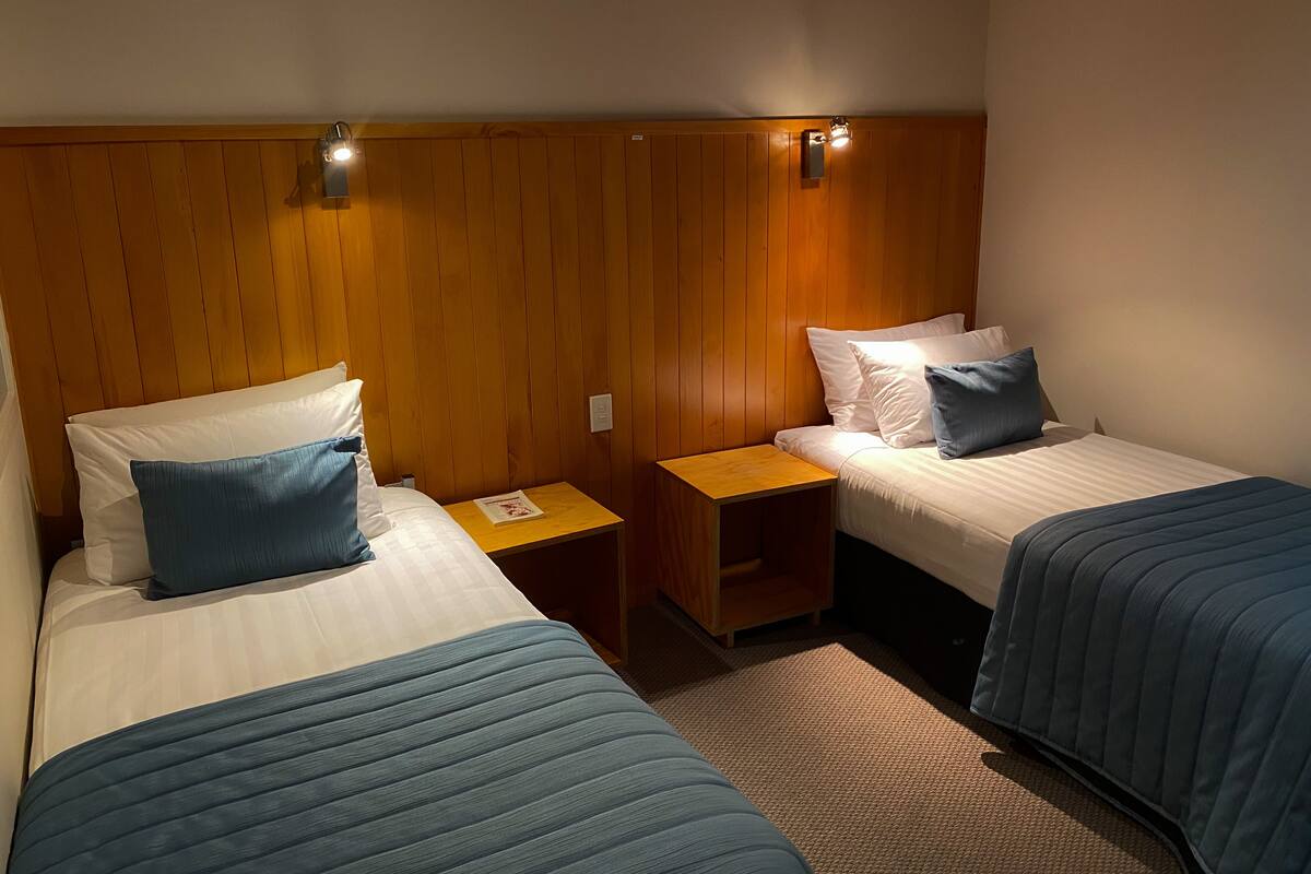 Quarto de hotel com duas camas de solteiro, paredes de painel de madeira, uma pequena mesa de cabeceira de madeira e luminária fixada na parede.