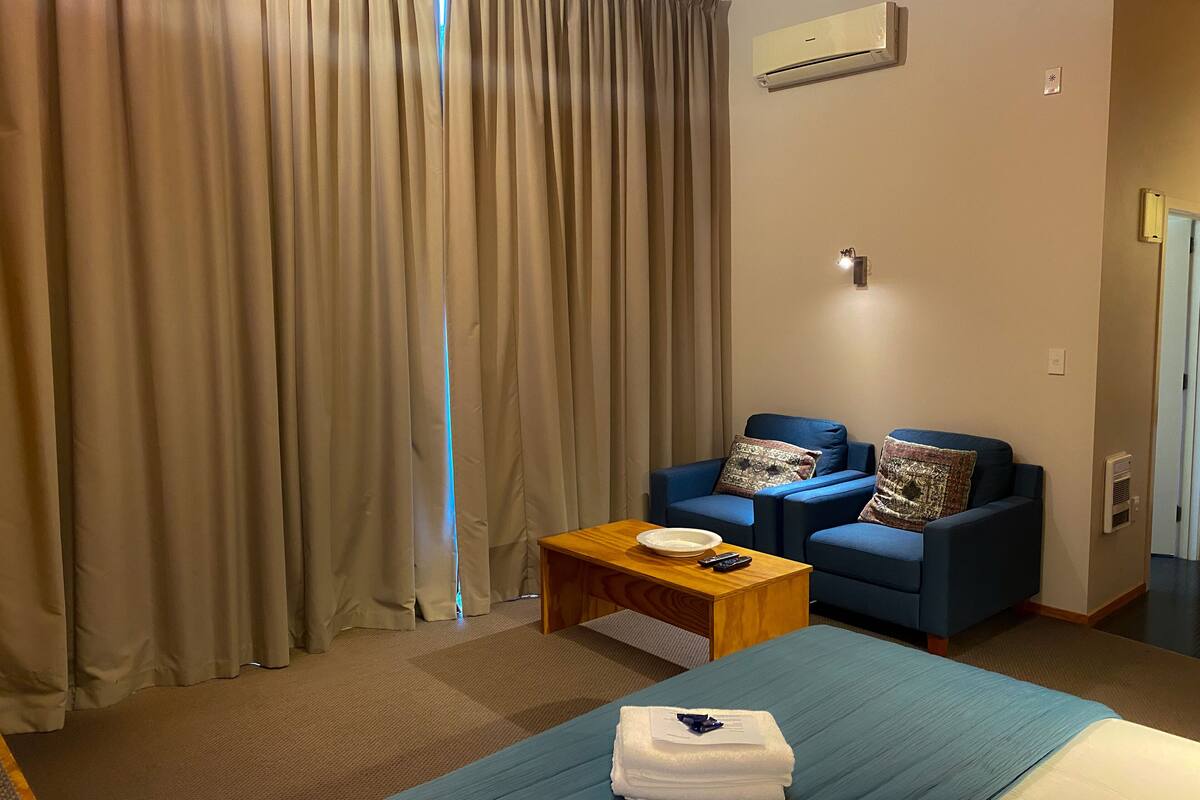 Quarto de hotel com uma cama, duas poltronas azuis, uma mesa de centro de madeira e cortinas bege.