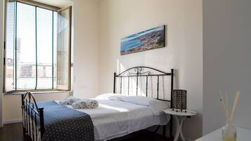 Uma cama bem arrumada com toalhas enroladas, uma pequena mesa de cabeceira com abajur e um quadro de paisagem costeira na parede.