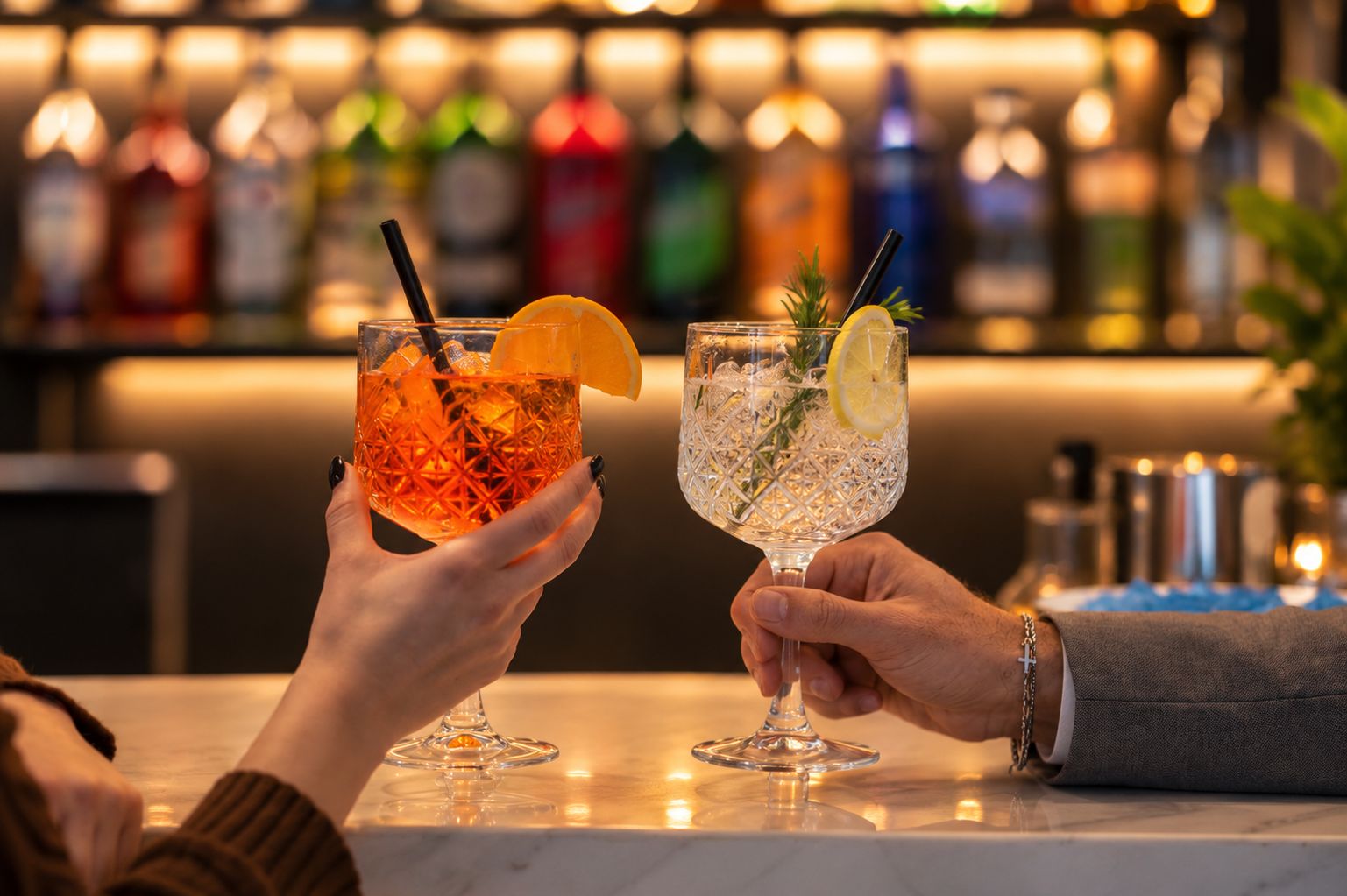 Duas mãos segurando copos de coquetel com guarnições, um bar com garrafas ao fundo.