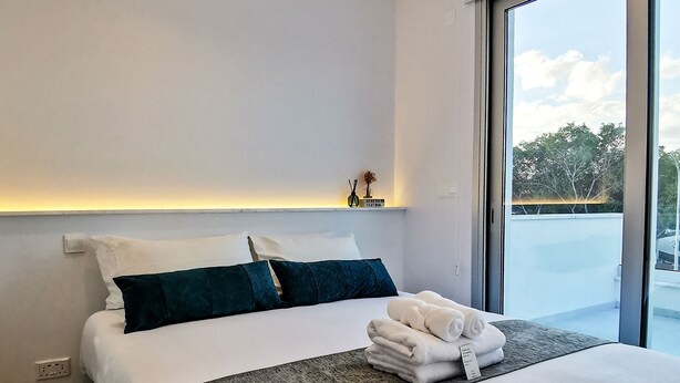 Quarto de hotel moderno com uma cama grande, vista para o exterior e uma pequena estante com objetos decorativos.