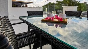 Uma mesa com tampo de vidro, cadeiras de vime, uma bandeja com flores cor-de-rosa e vista para o céu e árvores.