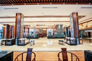 Um hall amplo com área de estar central, balcão de recepção e um espelho grande que reflete o ambiente.
