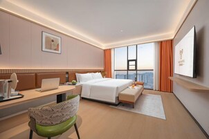 Quarto de hotel com cama, escrivaninha, cadeira e uma janela grande com cortinas.