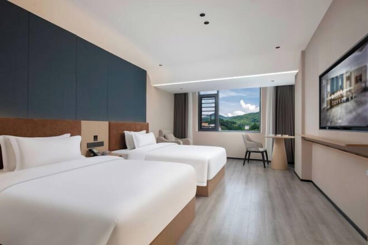 Quarto de hotel com duas camas, uma escrivaninha, uma cadeira e uma janela ampla com vista.