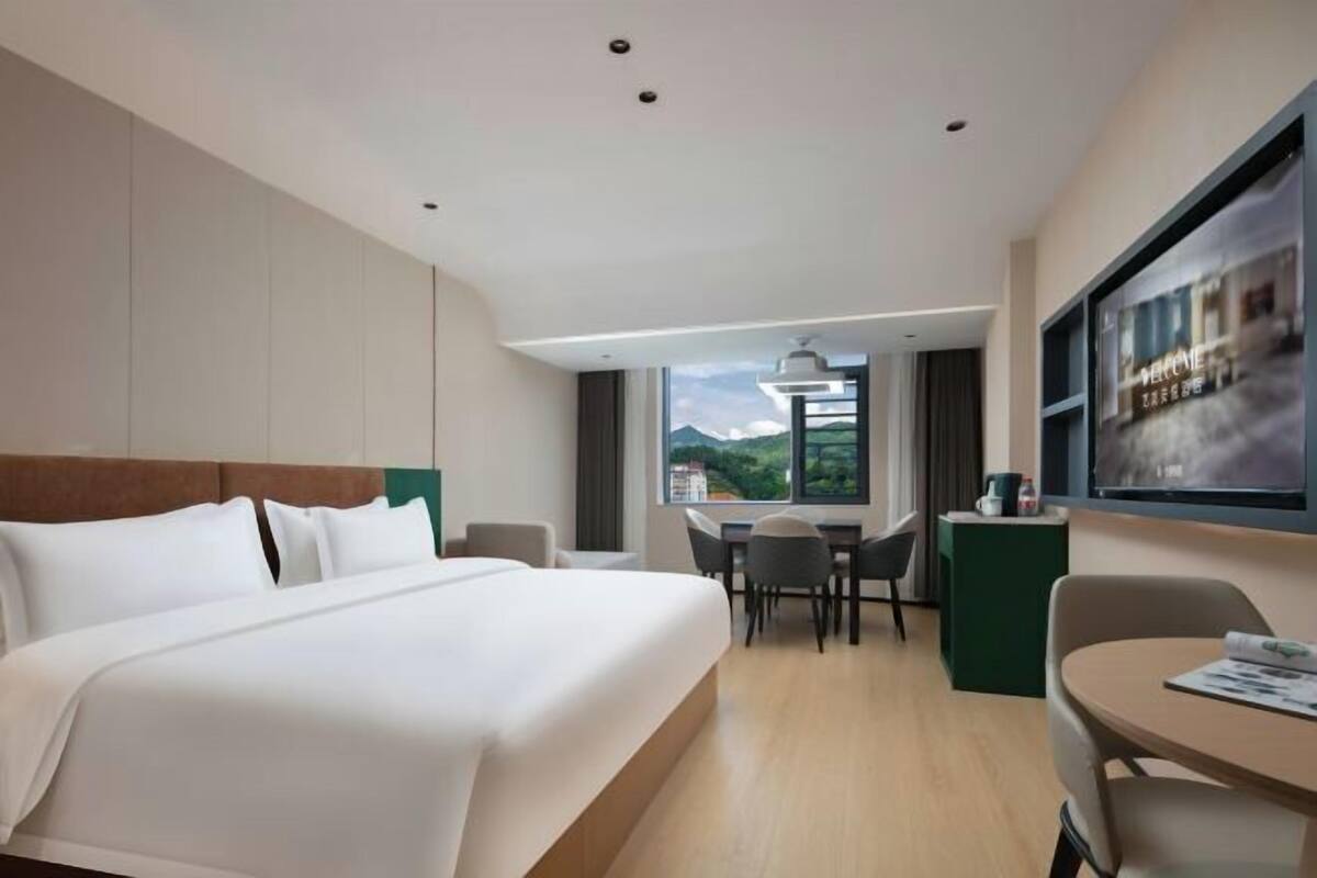 Quarto de hotel com uma cama grande, uma escrivaninha com cadeiras, vista para montanhas e uma televisão na parede.