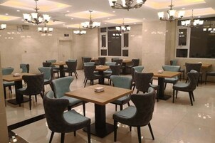 Uma sala de jantar de hotel com várias mesas e cadeiras, lustres e uma grande janela.