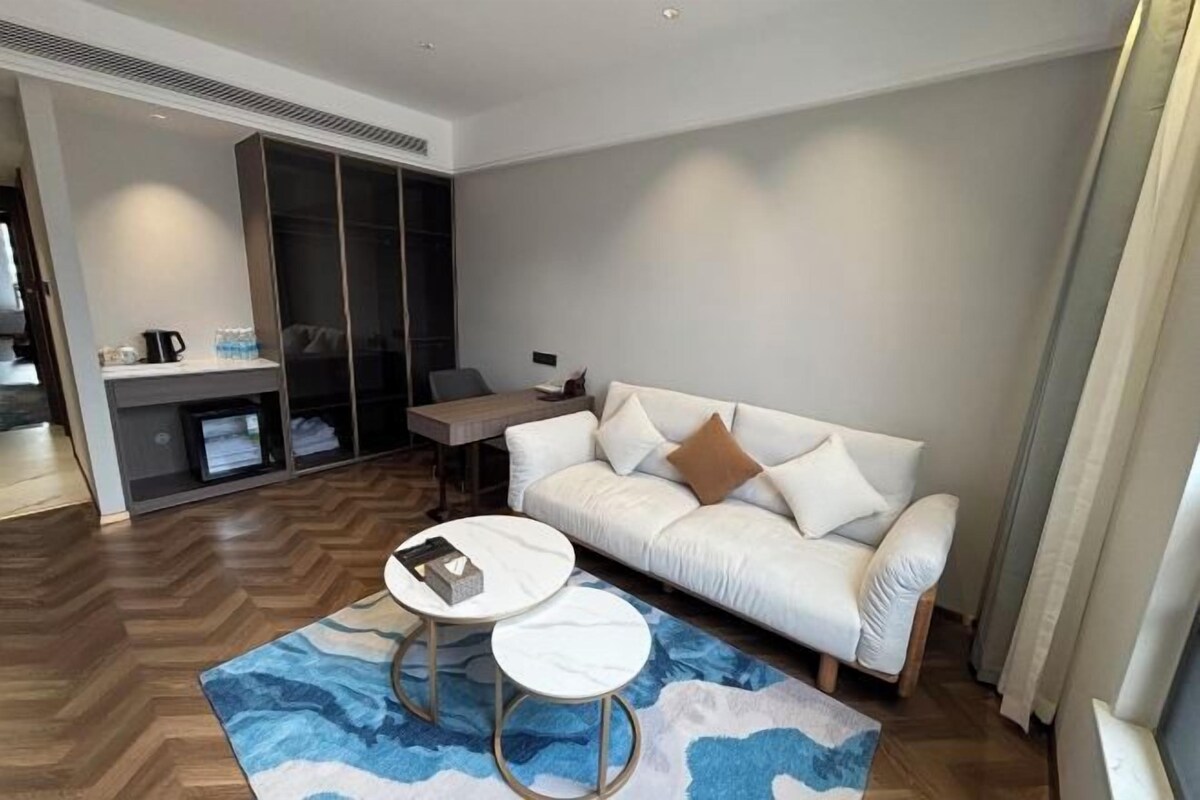 Quarto de hotel moderno com sofá branco, uma mesa redonda pequena e piso de madeira em padrão herringbone.