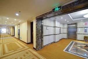 Corredor de hotel com balcão de recepção, elevadores e piso coberto por carpete.