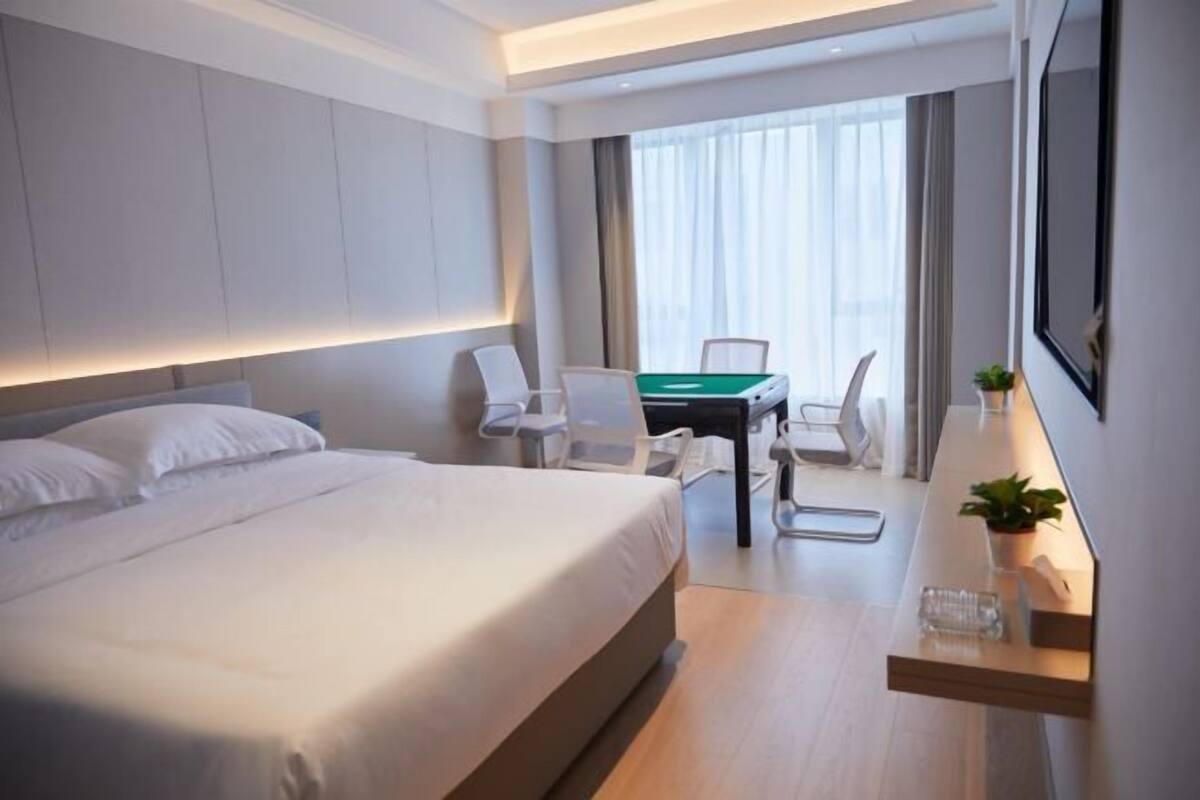 Quarto de hotel moderno com uma cama grande, uma mesa com um jogo de tabuleiro verde, uma cadeira e uma pequena planta.
