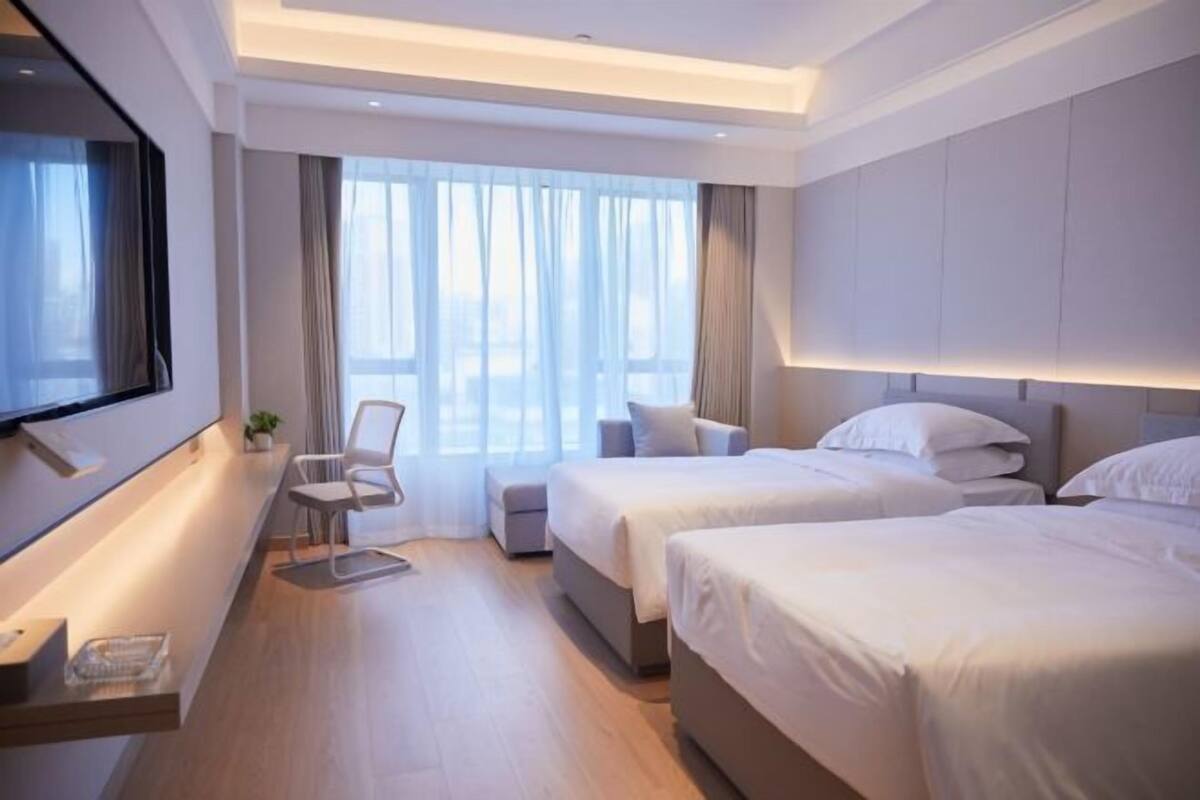 Quarto de hotel moderno com duas camas, uma escrivaninha com cadeira e uma janela grande com cortinas.