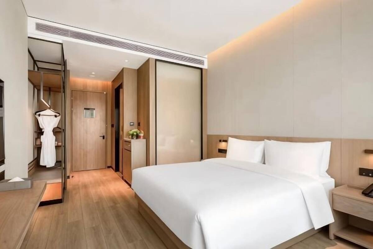 Quarto de hotel moderno com uma cama grande, mesa de cabeceira, guarda-roupa e banheiro visível por uma porta aberta.