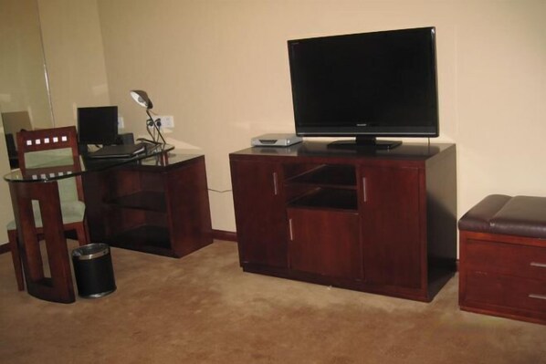 Um quarto com uma escrivaninha de madeira, uma televisão em um suporte, uma cadeira e uma lixeira.