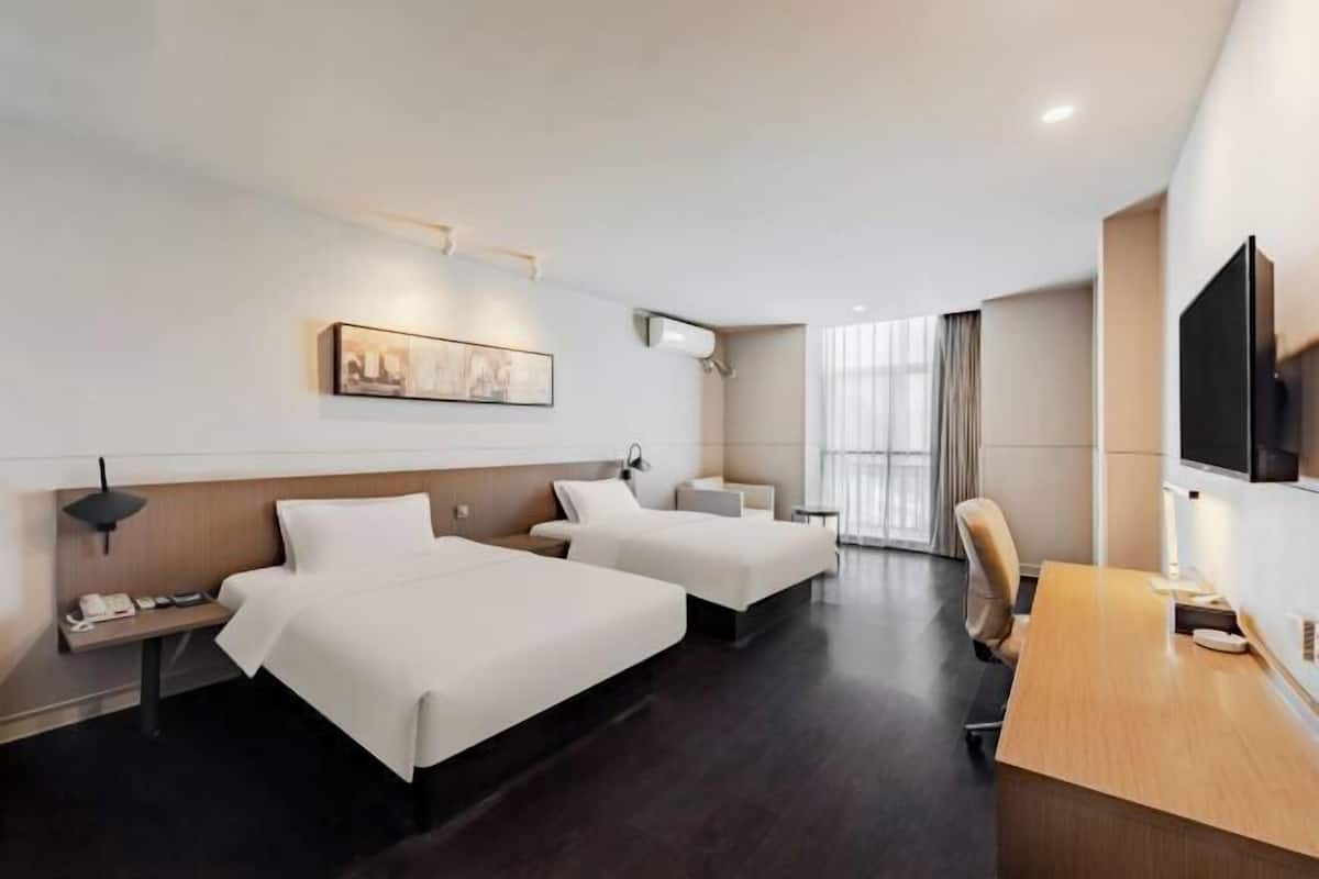 Quarto de hotel com duas camas, uma escrivaninha, uma cadeira, televisão de tela plana, um quadro na parede e uma janela com cortinas.
