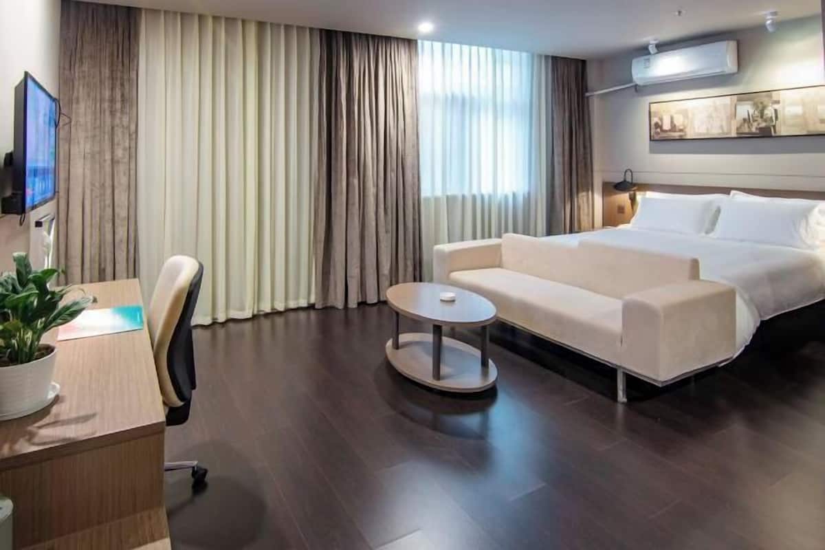 Quarto de hotel com cama, sofá, mesa pequena, escrivaninha, televisão e planta em vaso.
