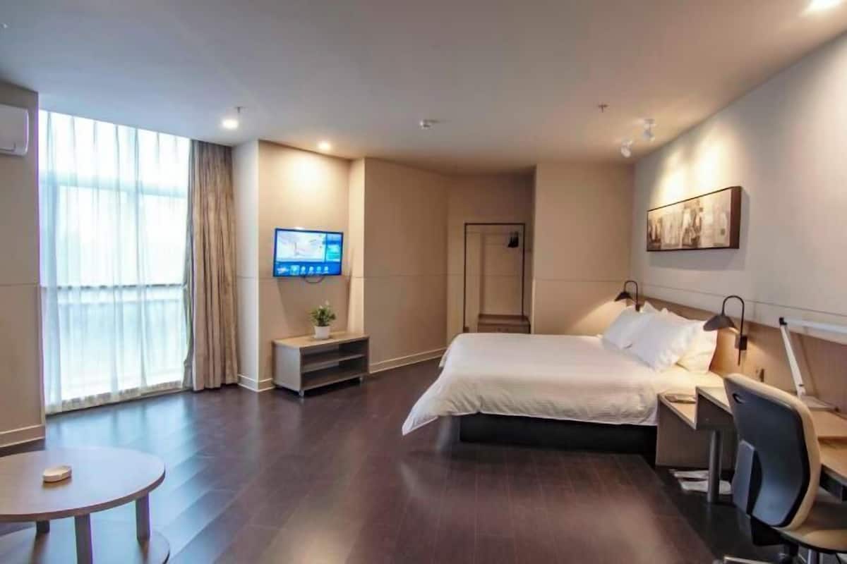 Quarto de hotel moderno com cama grande, mesa de cabeceira, cadeira, TV e pequena mesa.
