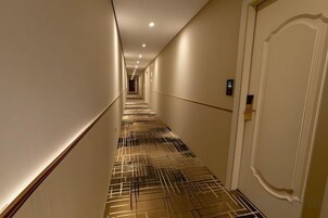 Corredor de hotel com piso estampado, uma porta à direita e um lustre no teto.